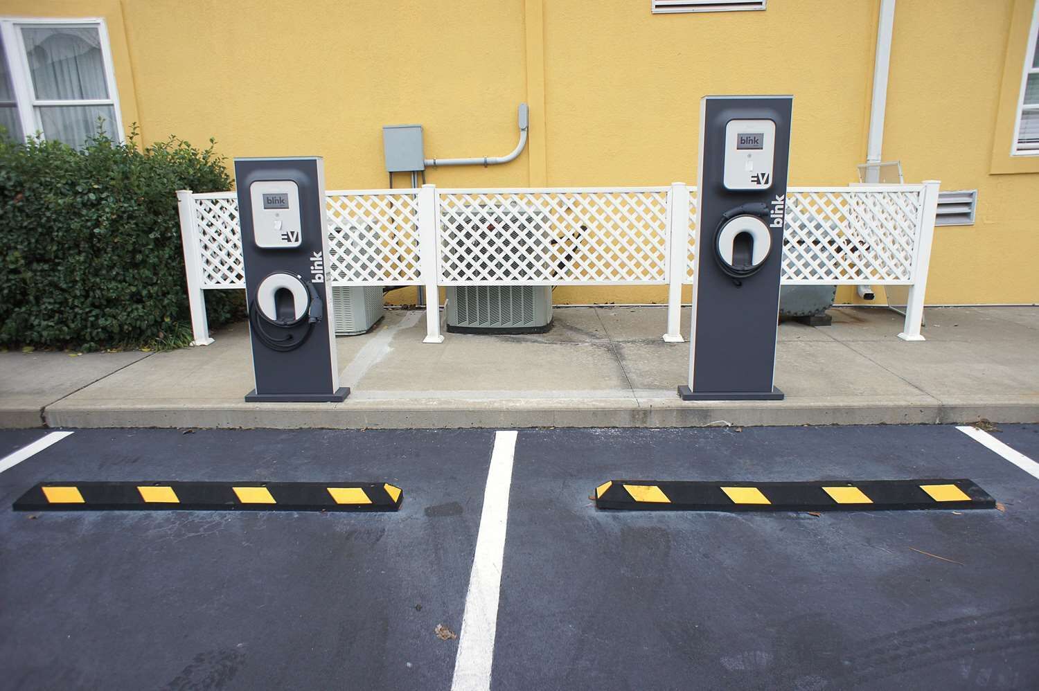 Duas estações de carregamento para veículos elétricos, com uma cerca de grade branca e um edifício amarelo ao fundo.