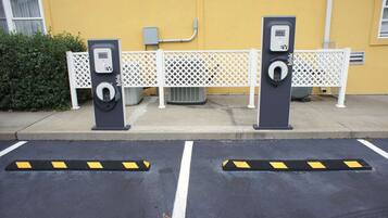 Duas estações de carregamento para veículos elétricos, com uma cerca de grade branca e um edifício amarelo ao fundo.