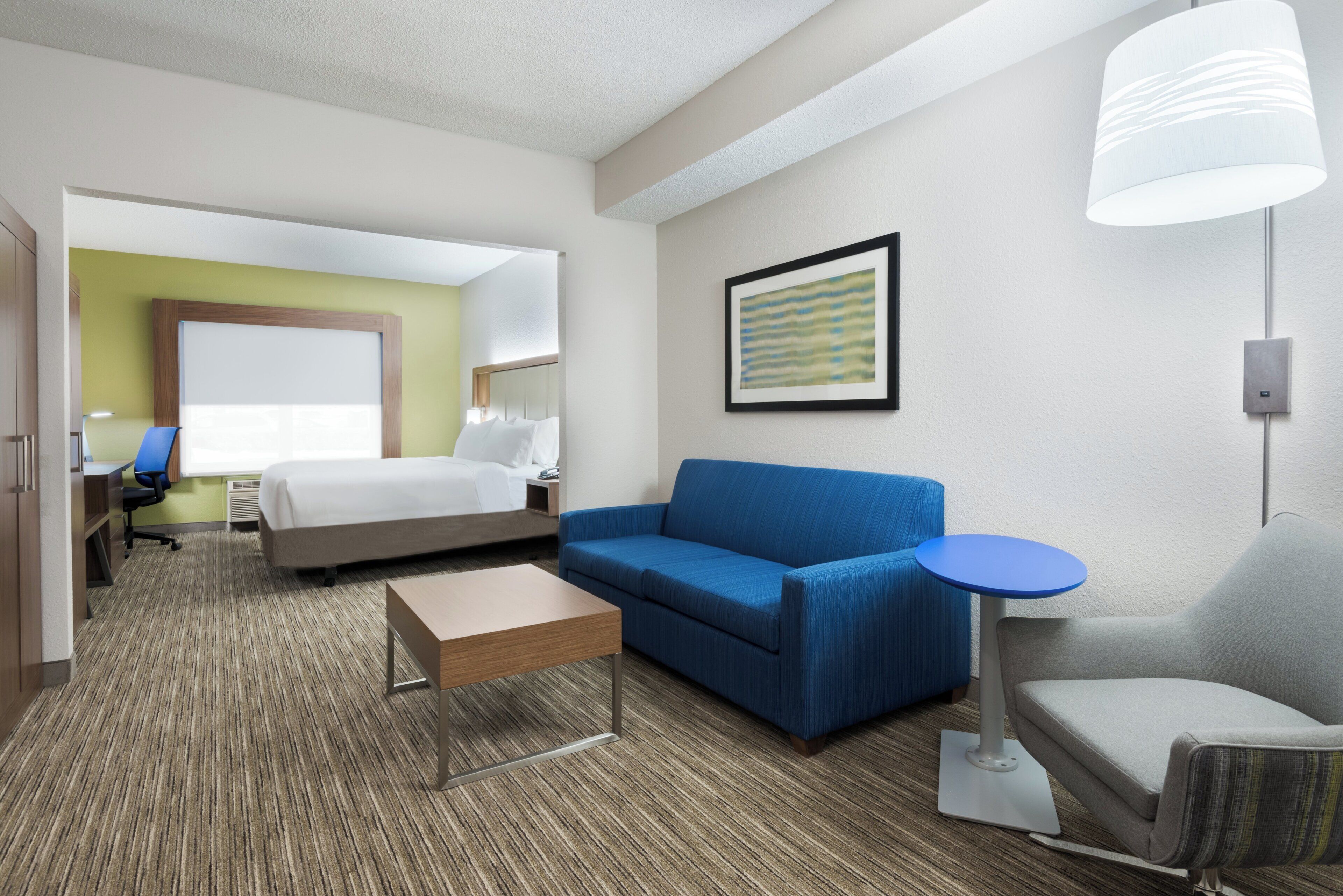Quarto de hotel com cama, sofá azul, mesa pequena, cadeira, escrivaninha e abajur.