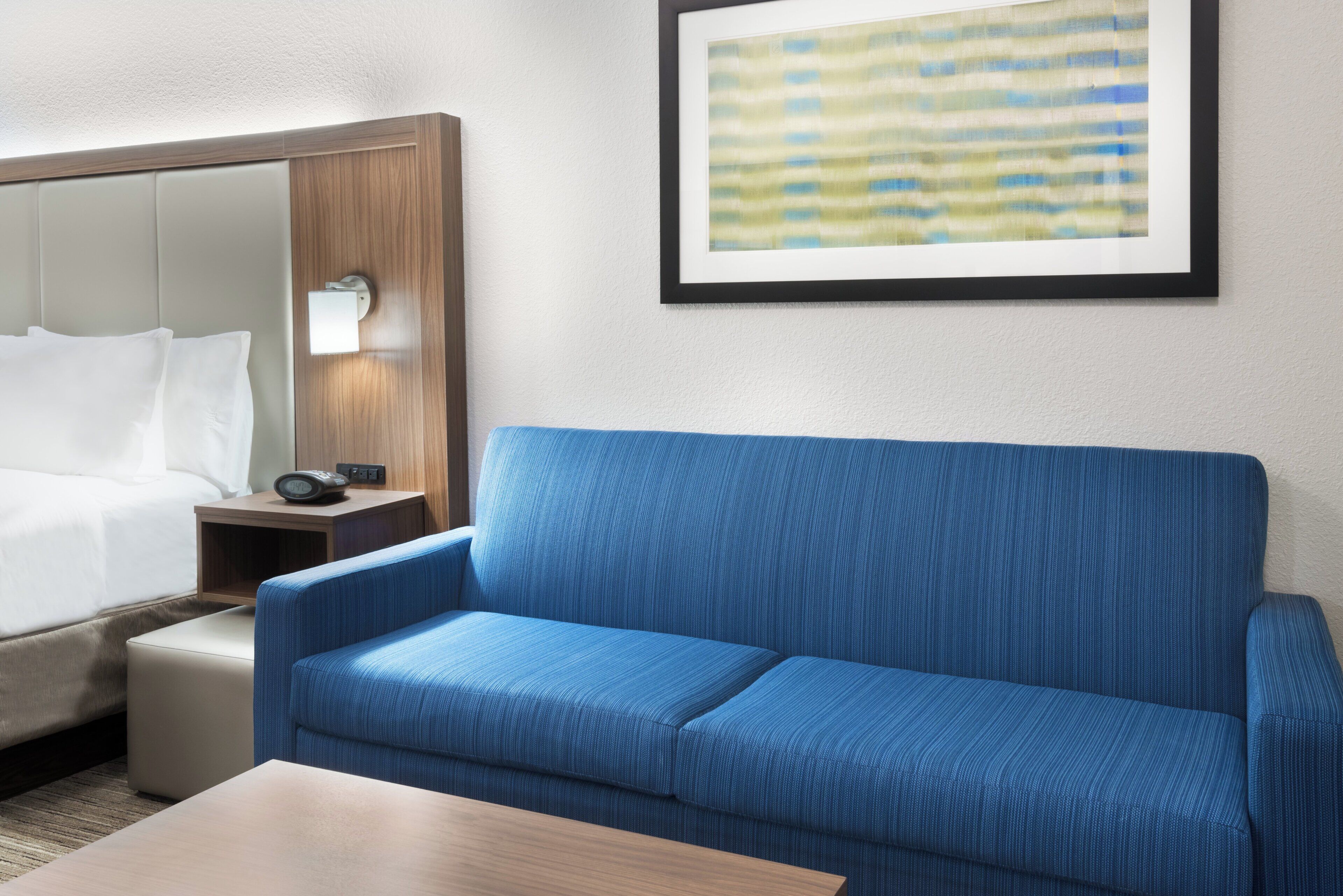 Quarto de hotel com cama, sofá azul, mesa de cabeceira de madeira com abajur e uma pintura abstrata emoldurada na parede.