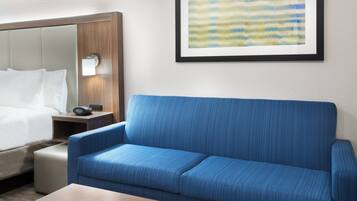 Quarto de hotel com cama, sofá azul, mesa de cabeceira de madeira com abajur e uma pintura abstrata emoldurada na parede.