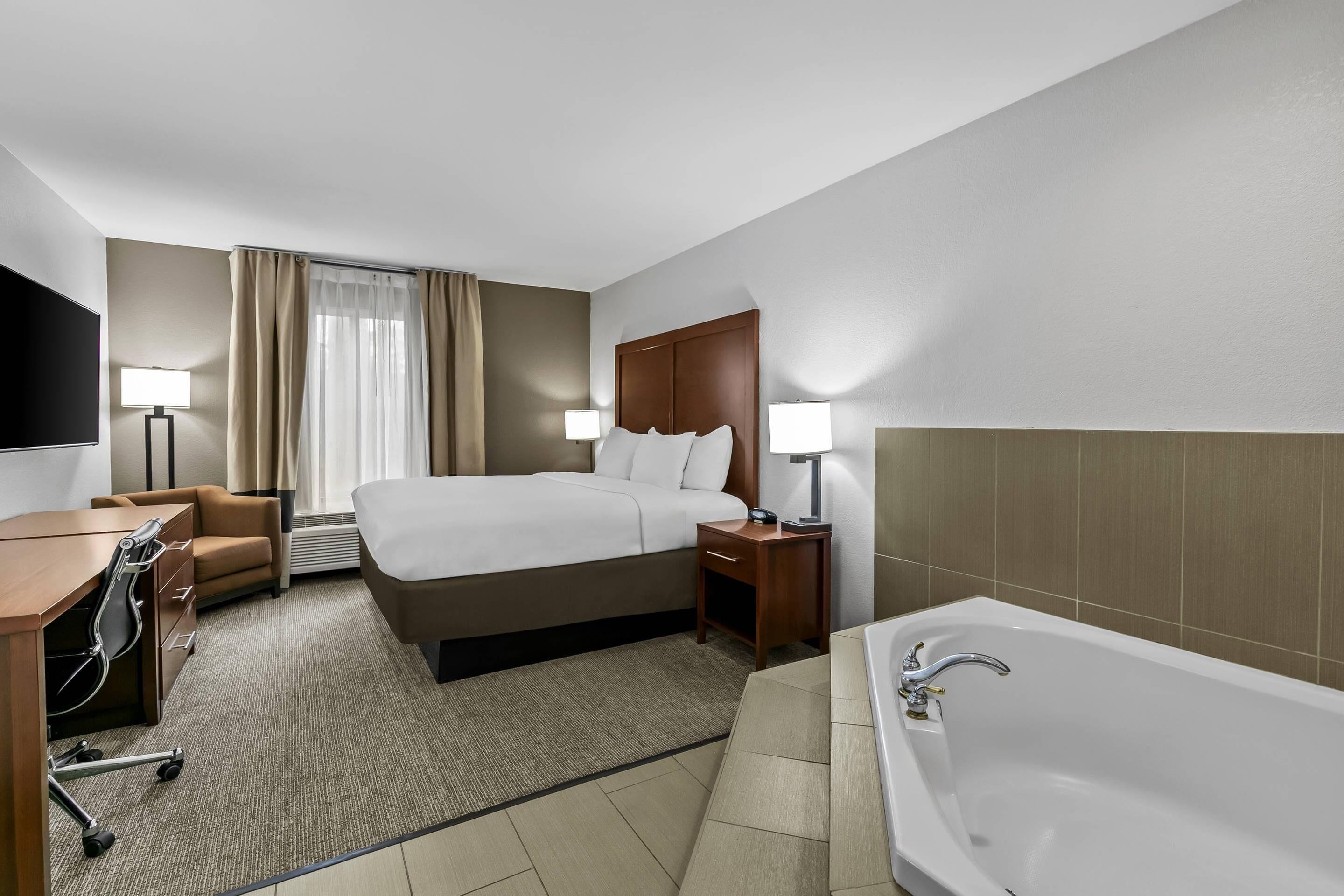 Quarto de hotel com cama, escrivaninha, cadeira, abajur e banheira.