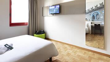 Quarto de hotel com cama, televisão fixada na parede, escrivaninha com cadeira e um espelho grande que reflete o quarto.