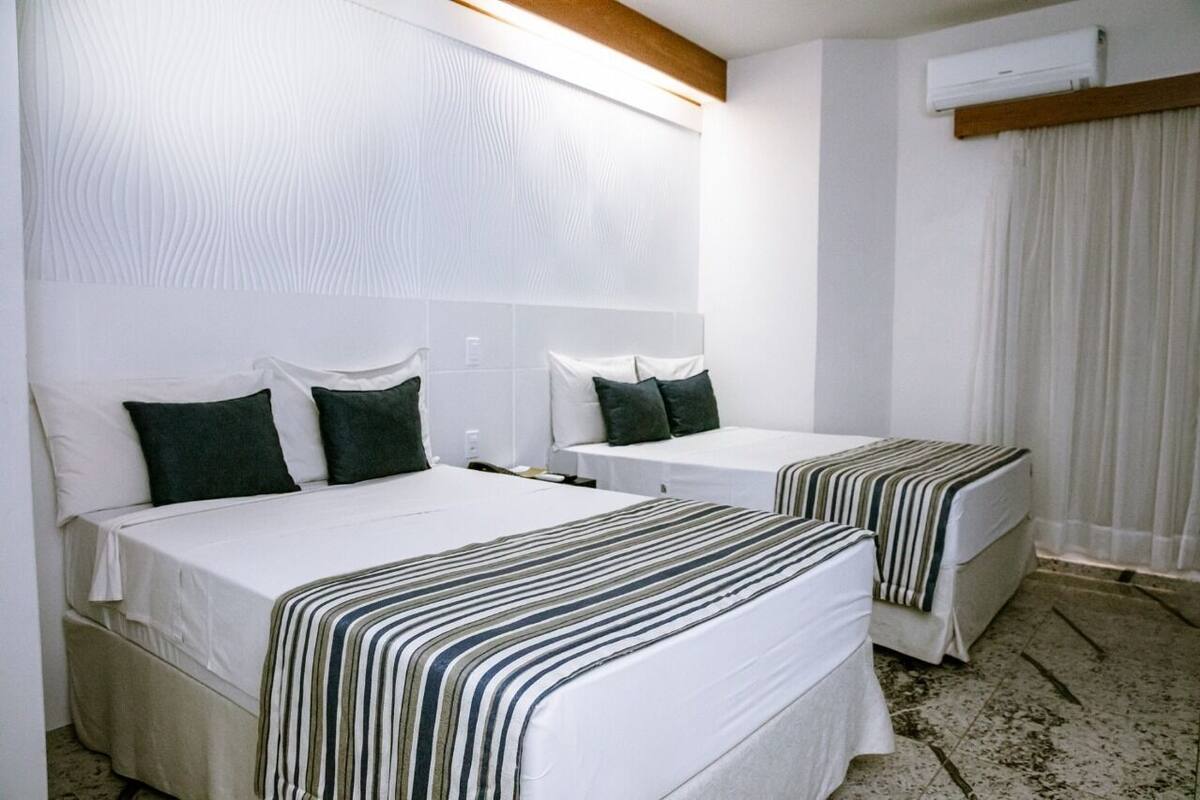 Quarto de hotel com duas camas, cada uma com roupa de cama branca e cobertores listrados. Possui cabeceira de madeira, parede branca e janela com cortinas brancas.