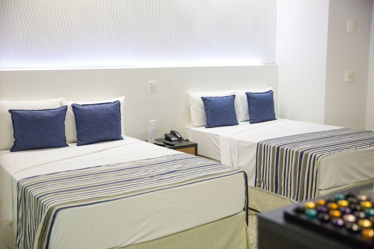 Quarto de hotel com duas camas, cada uma com colcha listrada e almofadas azuis. Há uma mesa de cabeceira com telefone e abajur entre as camas.