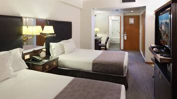 Quarto de hotel com duas camas, uma escrivaninha com telefone, televisão e banheiro visível por uma porta aberta.