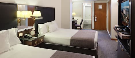 Quarto de hotel com duas camas, uma escrivaninha com telefone, televisão e banheiro visível por uma porta aberta.