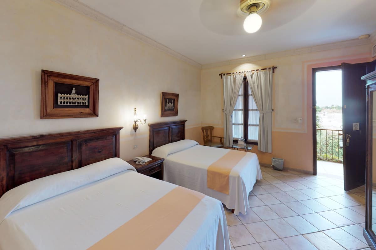 Quarto de hotel com duas camas, uma mesinha, uma cadeira, uma janela com cortinas e uma porta para o exterior.