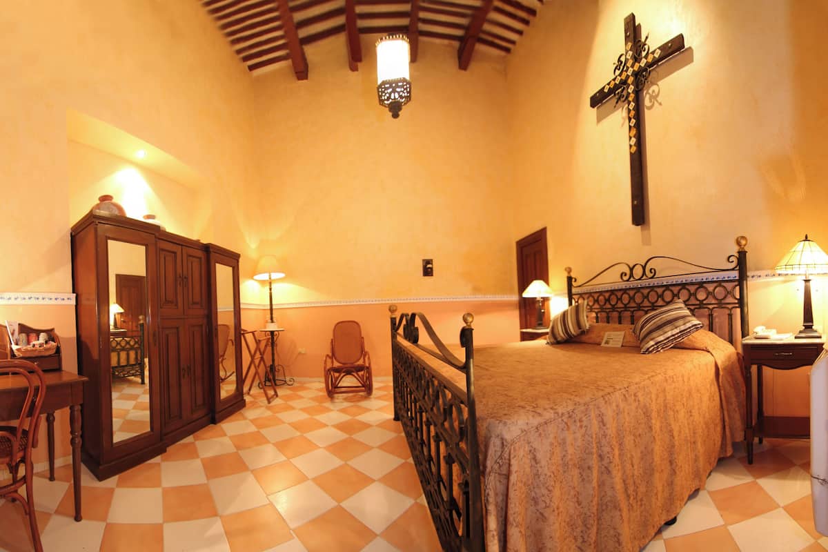 Quarto de hotel com piso xadrez, cama com cabeceira de metal, guarda-roupa de madeira, uma cadeira, escrivaninha com abajur e uma cruz fixada na parede.