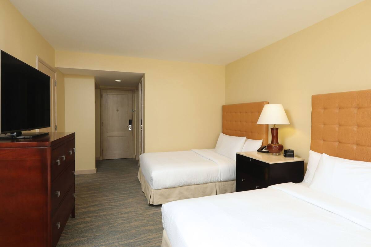Quarto de hotel com duas camas, uma TV em uma cômoda e um abajur em uma mesa de cabeceira.
