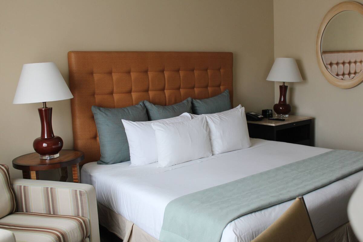 Quarto de hotel com uma cama grande, duas mesas de cabeceira com luminárias, uma poltrona listrada e um espelho na parede.