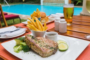Uma mesa de jantar com um prato de comida, incluindo um bife, batatas fritas e legumes, perto de uma piscina.