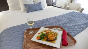 Cama bem arrumada com colcha azul, bandeja de madeira com prato de comida e um copo de vinho branco, e uma cadeira ao fundo.
