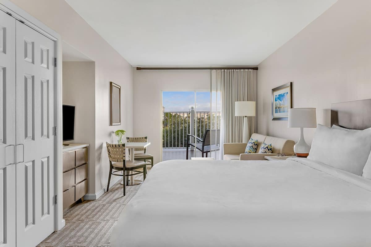 Quarto de hotel com uma cama grande, uma escrivaninha com cadeira, televisão, varanda com vista e janela com cortinas.