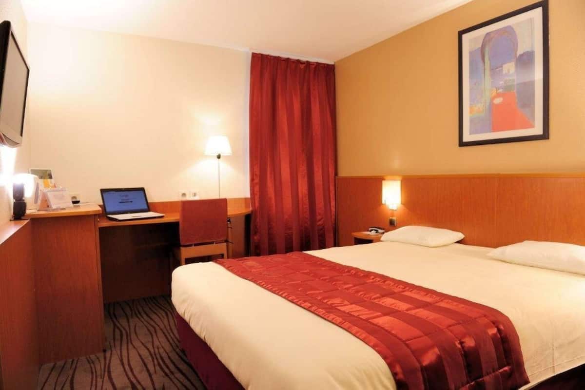 Quarto de hotel com cama, escrivaninha com laptop, cadeira, mesa de cabeceira e um quadro na parede.