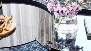 Uma mesa de centro redonda de vidro, com um vaso de flores e um prato de petiscos.