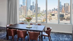 Quarto de hotel com uma grande janela com vista para a cidade, uma mesa de jantar com cadeiras e um vaso com flores.