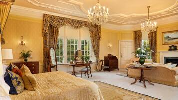 Um quarto luxuoso com uma cama grande, uma escrivaninha, uma poltrona e um sofá. Há um candelabro, uma lareira e uma janela ampla com cortinas.