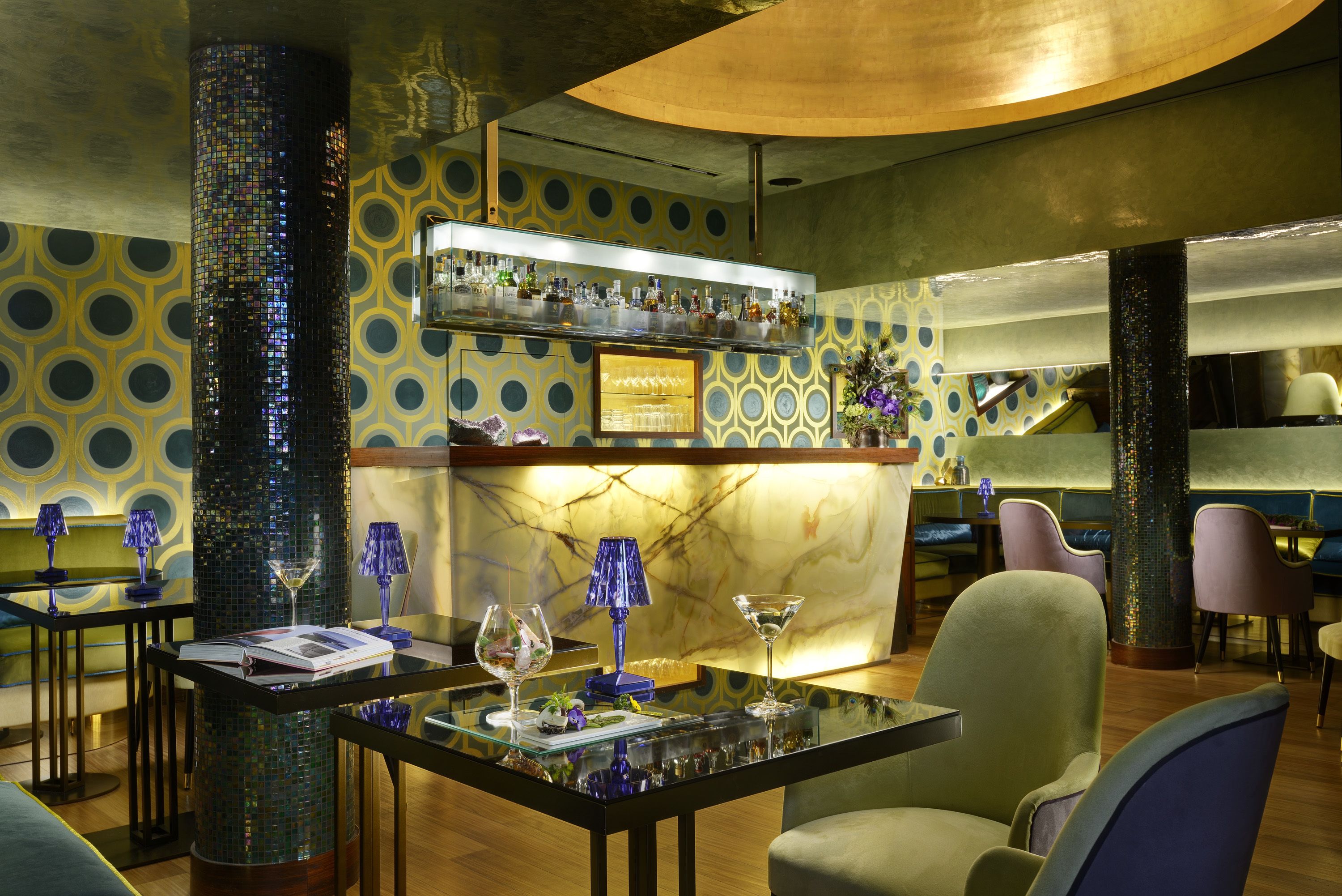 Uma área de bar com um pilar espelhado, banquetas e um espaço de estar com cadeiras acolchoadas.