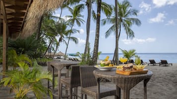 Uma área de refeições à beira-mar com móveis de vime, uma mesa posta com salada, hambúrguer, batatas fritas e bebidas, e palmeiras ao fundo.