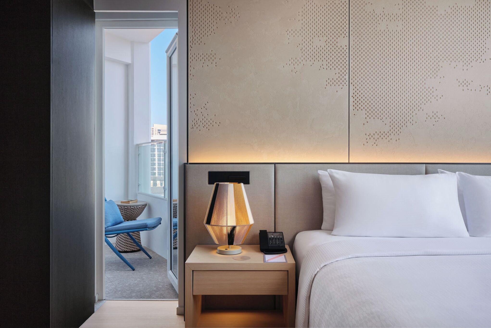 Quarto de hotel com cama, mesa de cabeceira com abajur, telefone e uma cadeira.