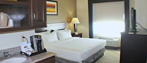 Quarto de hotel com cama, escrivaninha, luminária de mesa, cafeteira e uma janela grande.