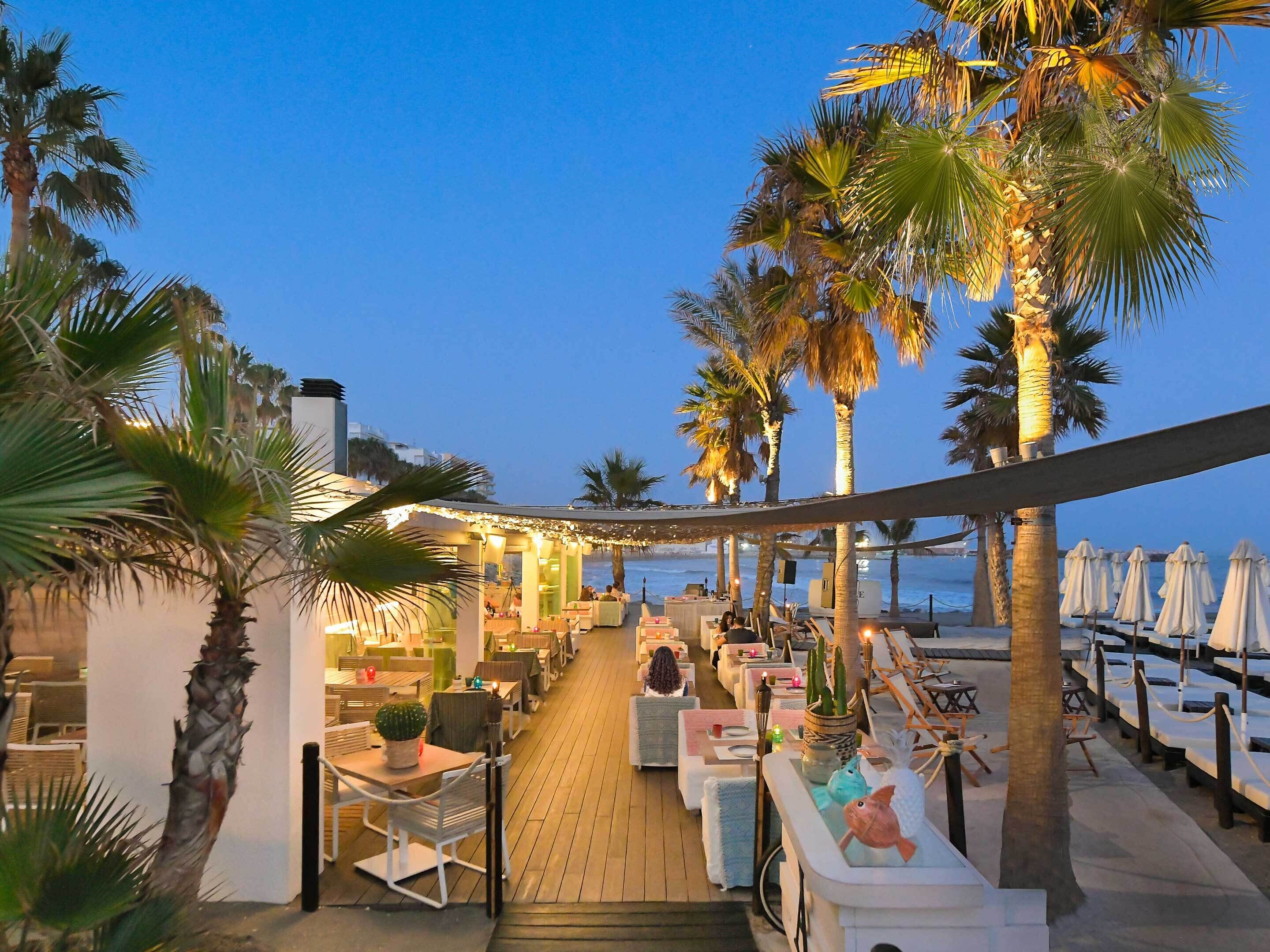 Um restaurante ao ar livre com mesas, cadeiras, palmeiras e vista para o mar.
