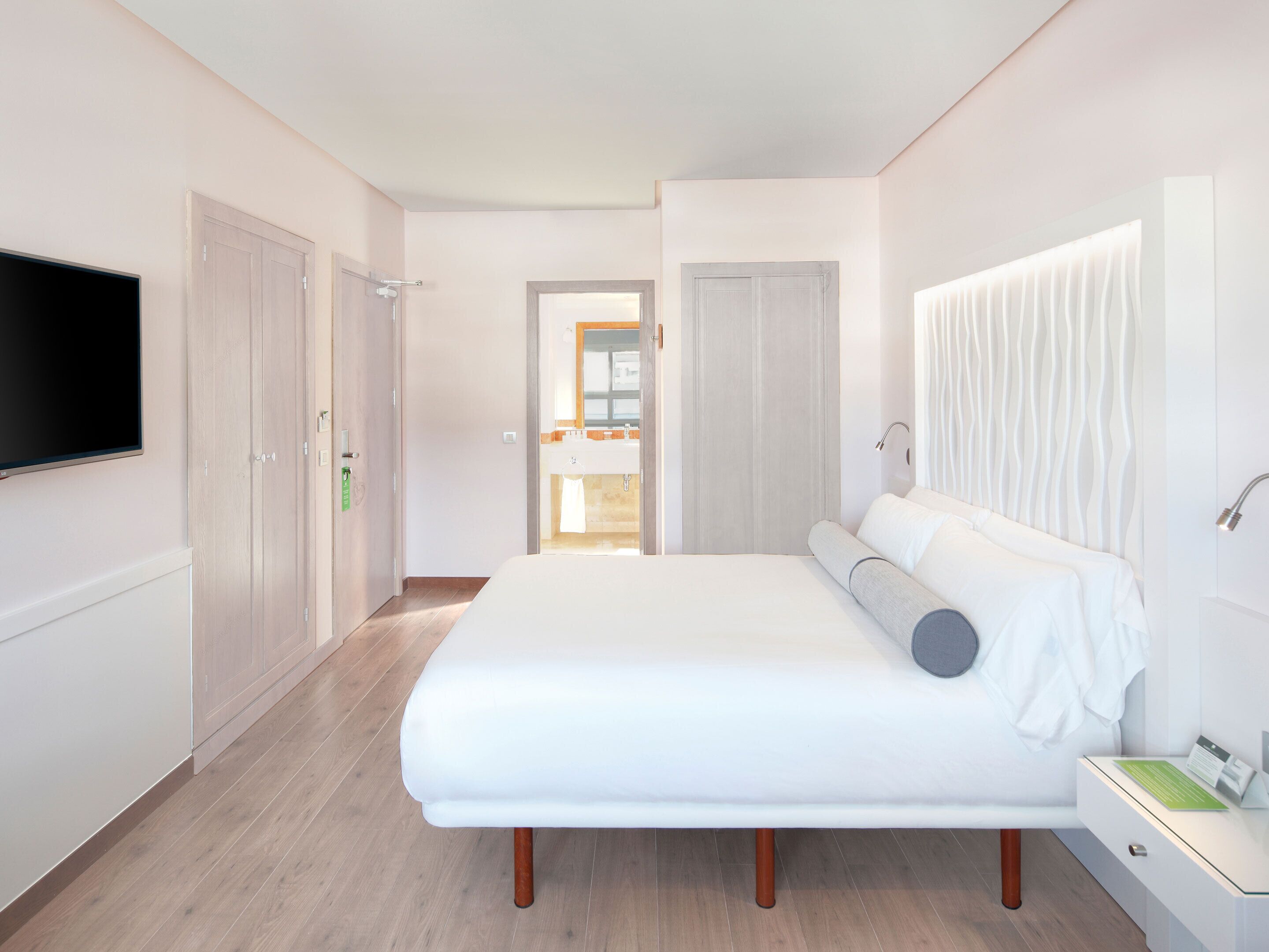 Quarto de hotel moderno com cama grande, televisão de tela plana, piso de madeira e vista para o banheiro.