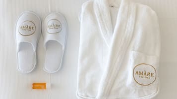 Um roupão branco e chinelos com a marca 'AMARE The One' bordada, colocados sobre uma superfície branca.