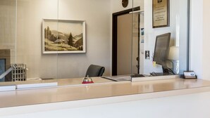 Uma receção com divisória de vidro, um quadro de paisagem emoldurado, um sino, uma luminária de mesa e uma placa na parede.