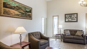 Uma sala de estar com sofá, poltrona e mesa de centro. Há duas luminárias, um quadro emoldurado e uma janela com cortinas.