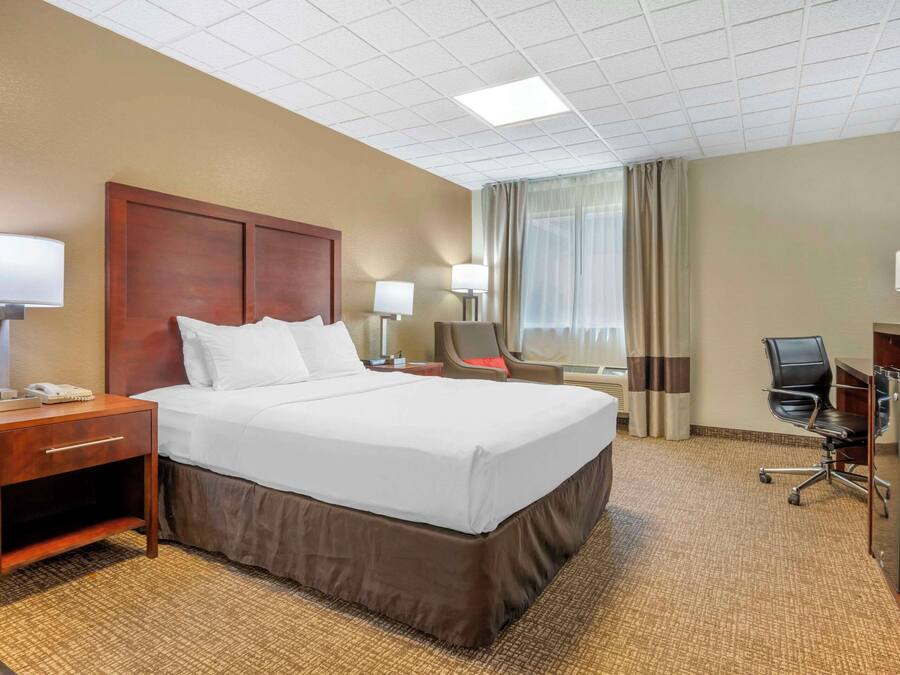 Quarto de hotel com uma cama grande, mesas de cabeceira com luminárias, uma escrivaninha com cadeira e uma janela com cortinas.