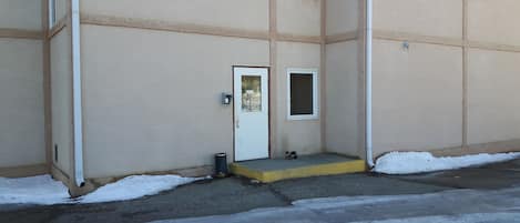 Entrada de um edifício com uma porta branca, uma pequena janela e um degrau amarelo que leva à porta.