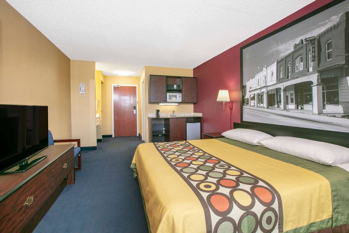 Quarto de hotel com uma cama, uma secretária com televisão, uma kitchenette e um quadro na parede.