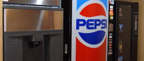 Uma área com máquinas de venda automática, incluindo um dispensador de Pepsi e uma geladeira de aço inoxidável.