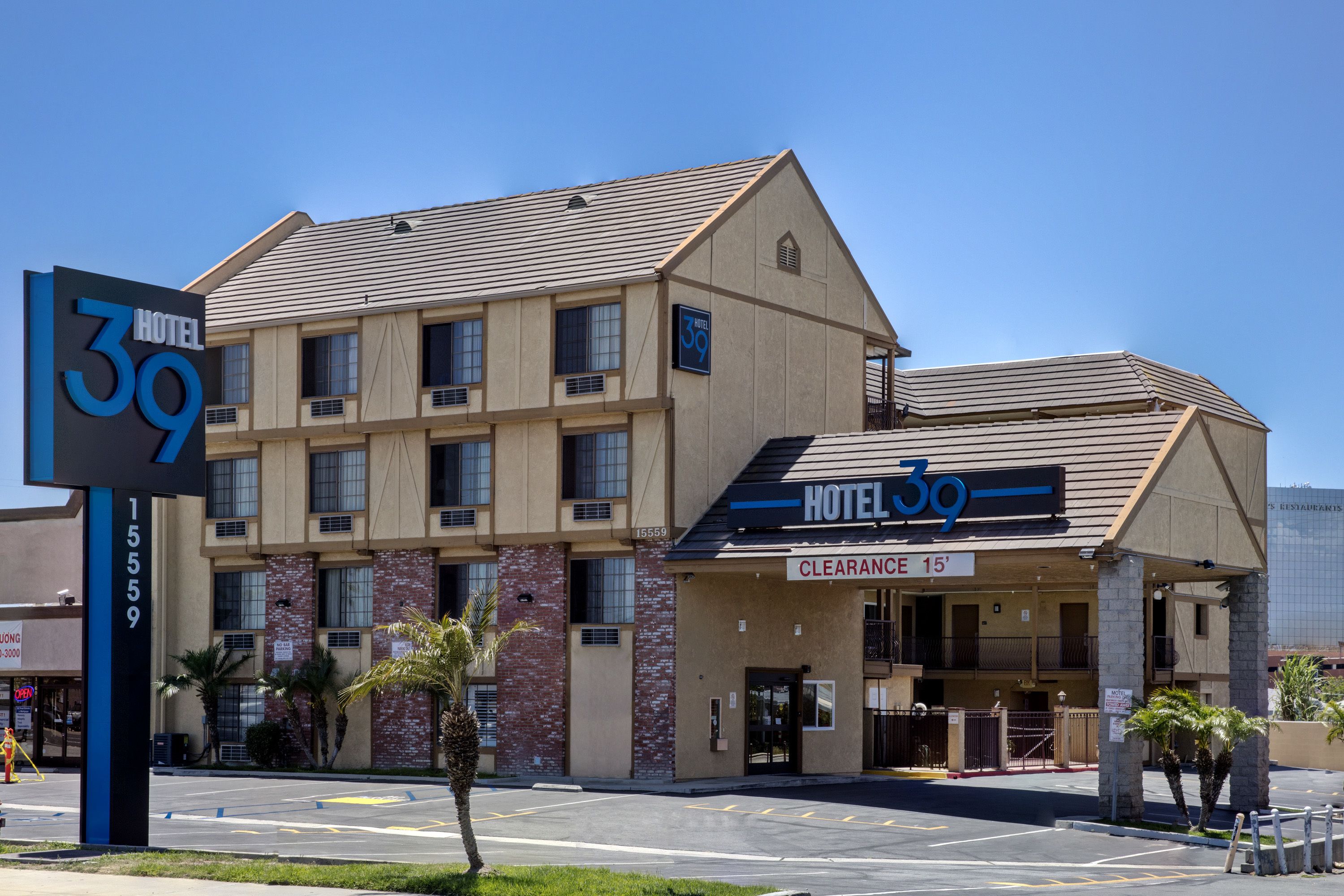 Um hotel de vários andares com uma placa de destaque 'Hotel 39', uma entrada visível e uma área de estacionamento.