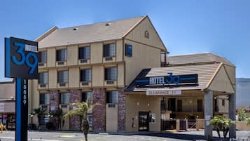 Um hotel de vários andares com uma placa de destaque 'Hotel 39', uma entrada visível e uma área de estacionamento.