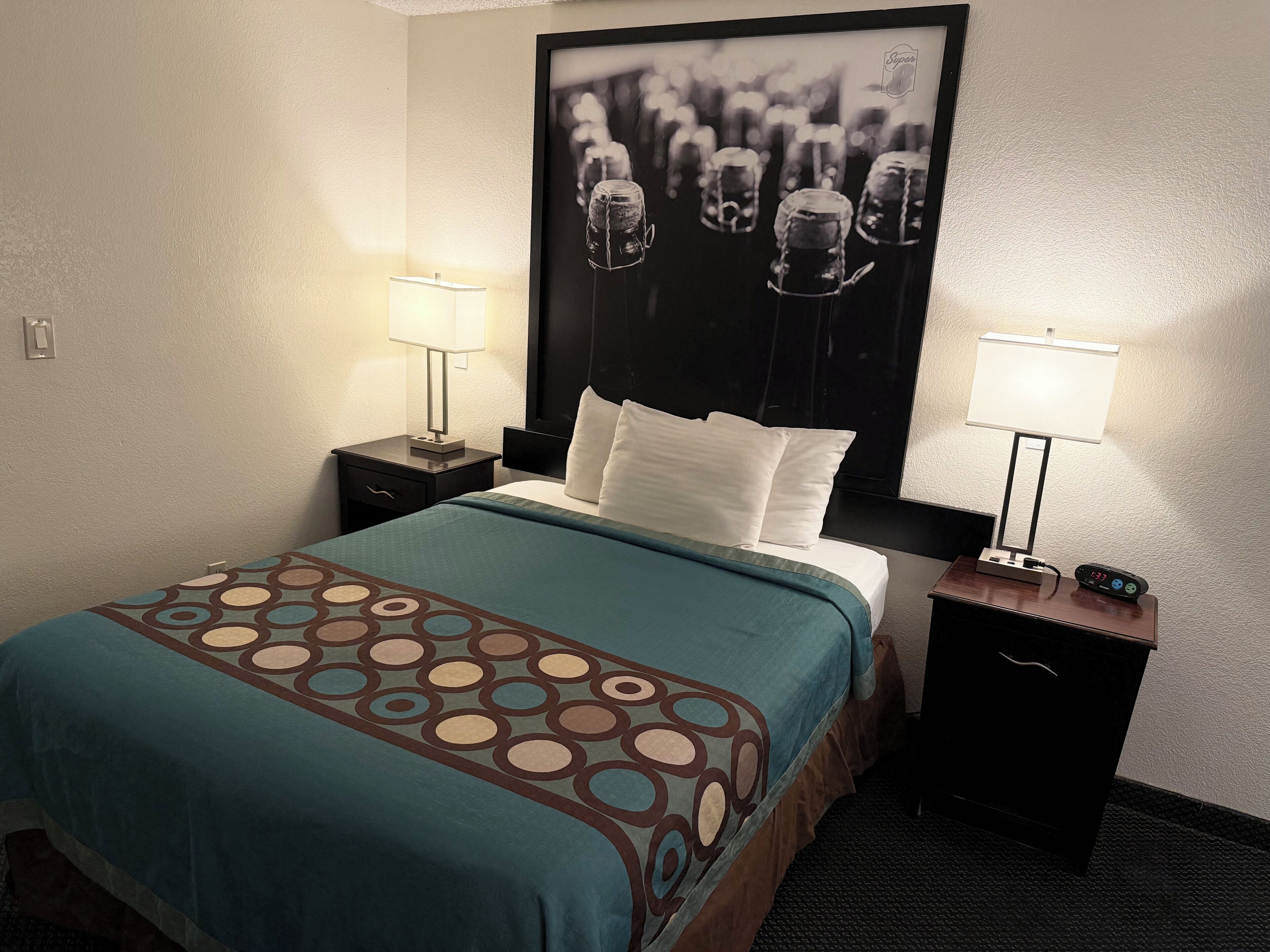 Quarto de hotel com uma cama, duas mesas de cabeceira, uma TV presa à parede e um cartaz grande sobre a cama.