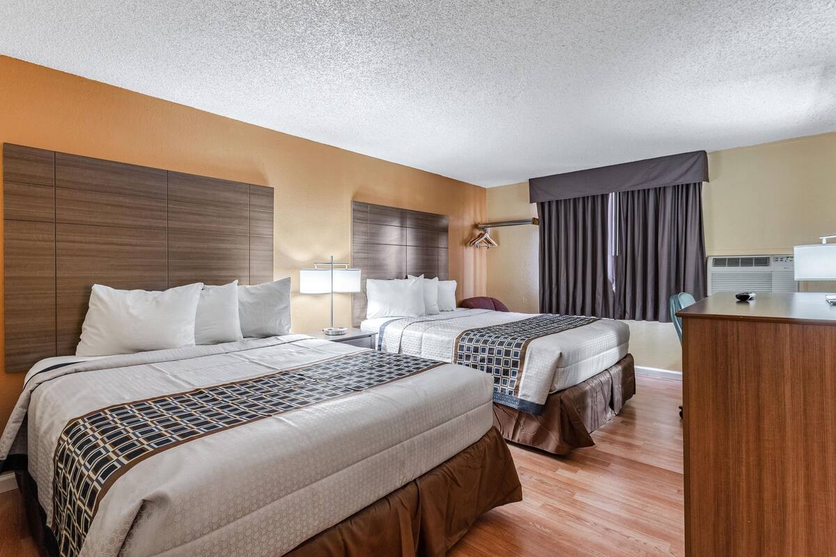 Quarto de hotel com duas camas, uma escrivaninha, cômoda e uma janela grande com cortinas.