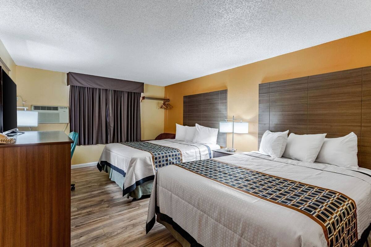 Quarto de hotel com duas camas, uma mesa, uma cadeira, uma televisão e uma janela com cortinas.