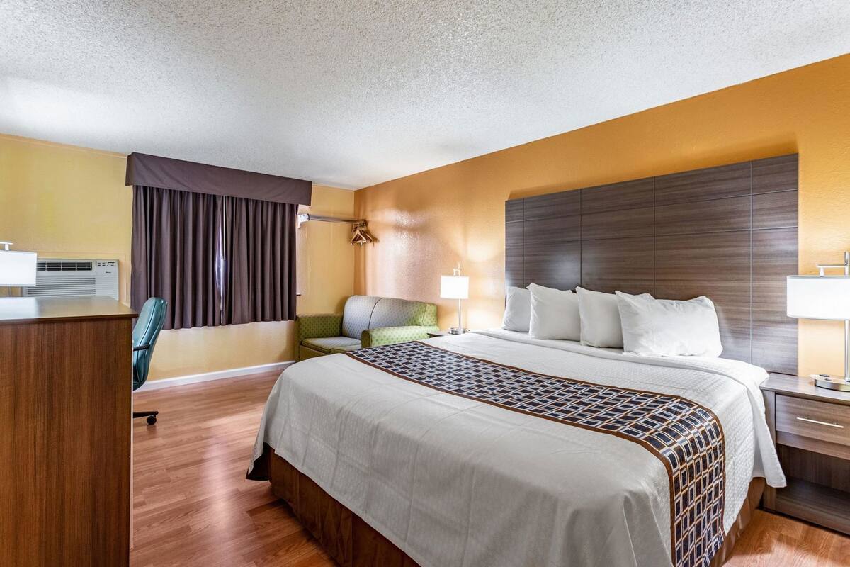 Quarto de hotel com cama, escrivaninha, cadeira, abajur e cortinas.
