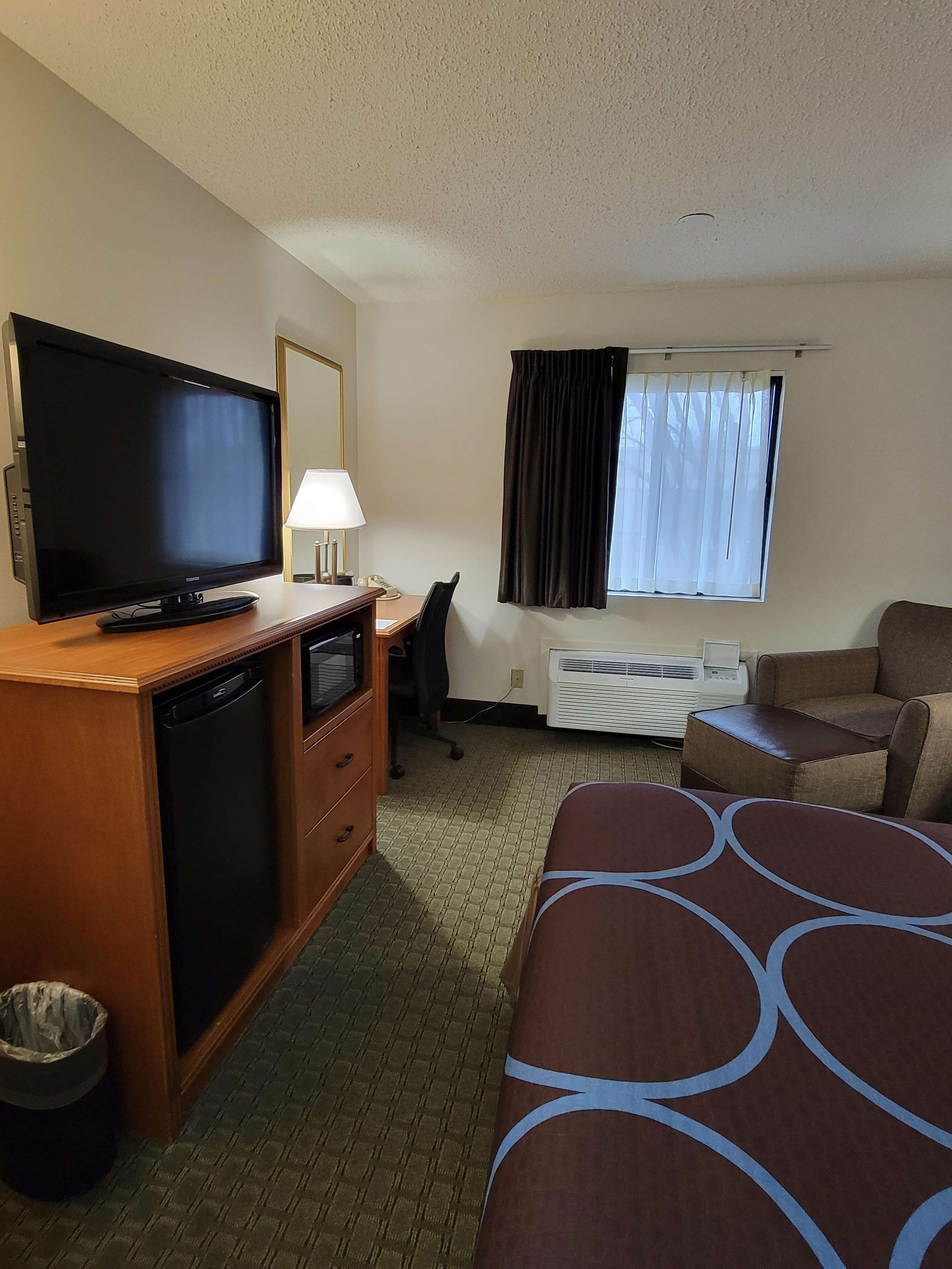 Quarto de hotel com cama, escrivaninha com televisão, candeeiro, cadeira, radiador e janela com cortinas.