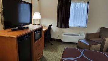 Quarto de hotel com cama, escrivaninha com televisão, candeeiro, cadeira, radiador e janela com cortinas.