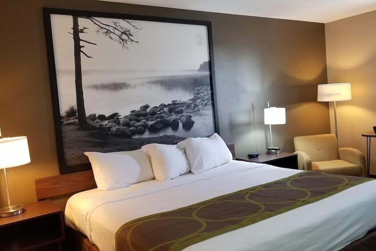 Quarto de hotel com uma cama grande, duas luminárias de cabeceira, uma mesa com cadeira e um quadro abstrato grande na parede.