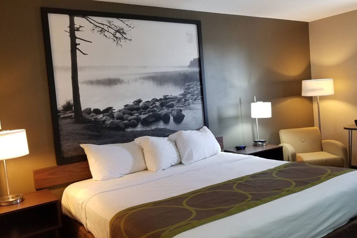 Quarto de hotel com cama, duas cadeiras, escrivaninha, abajur e uma grande moldura na parede.