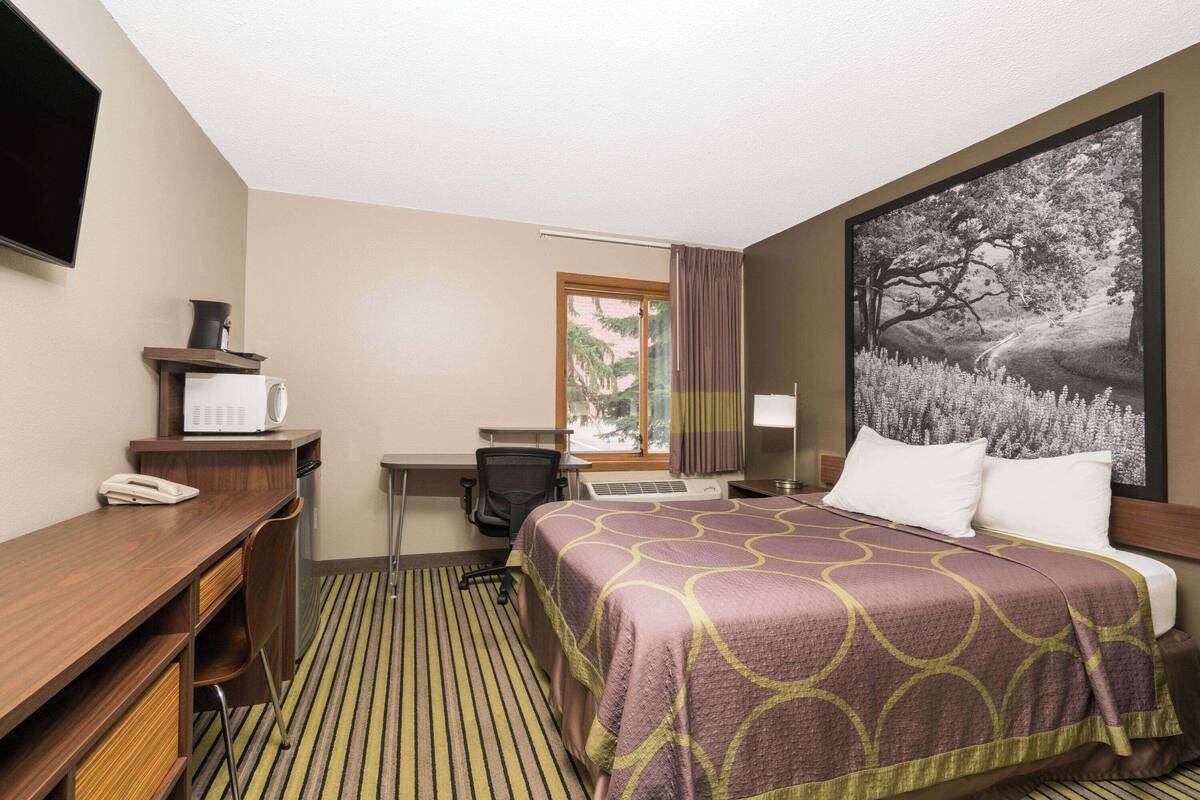 Quarto de hotel com cama, escrivaninha, cadeira e uma janela grande.