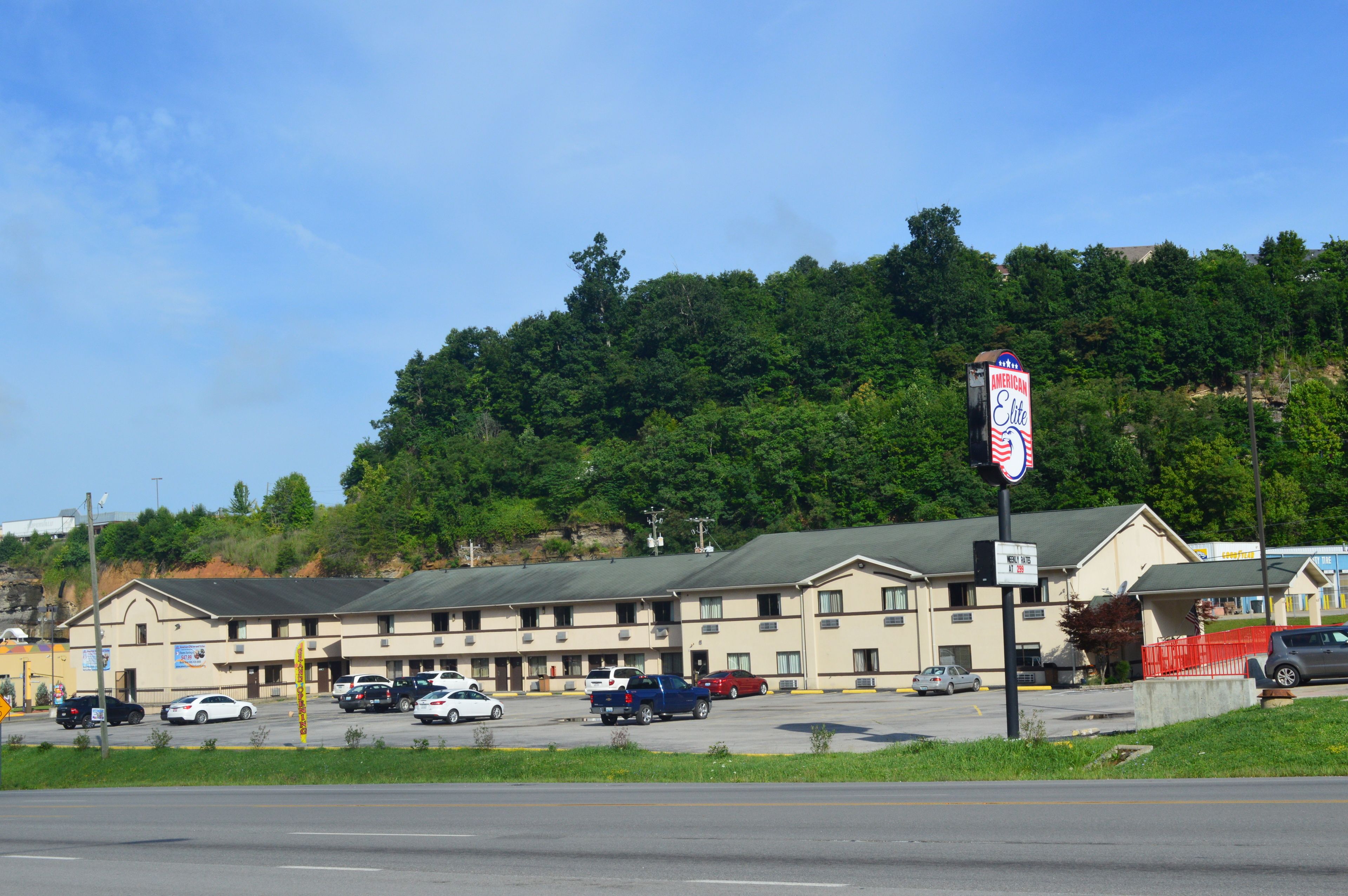 Um motel com várias salas, estacionamento e uma colina arborizada ao fundo.