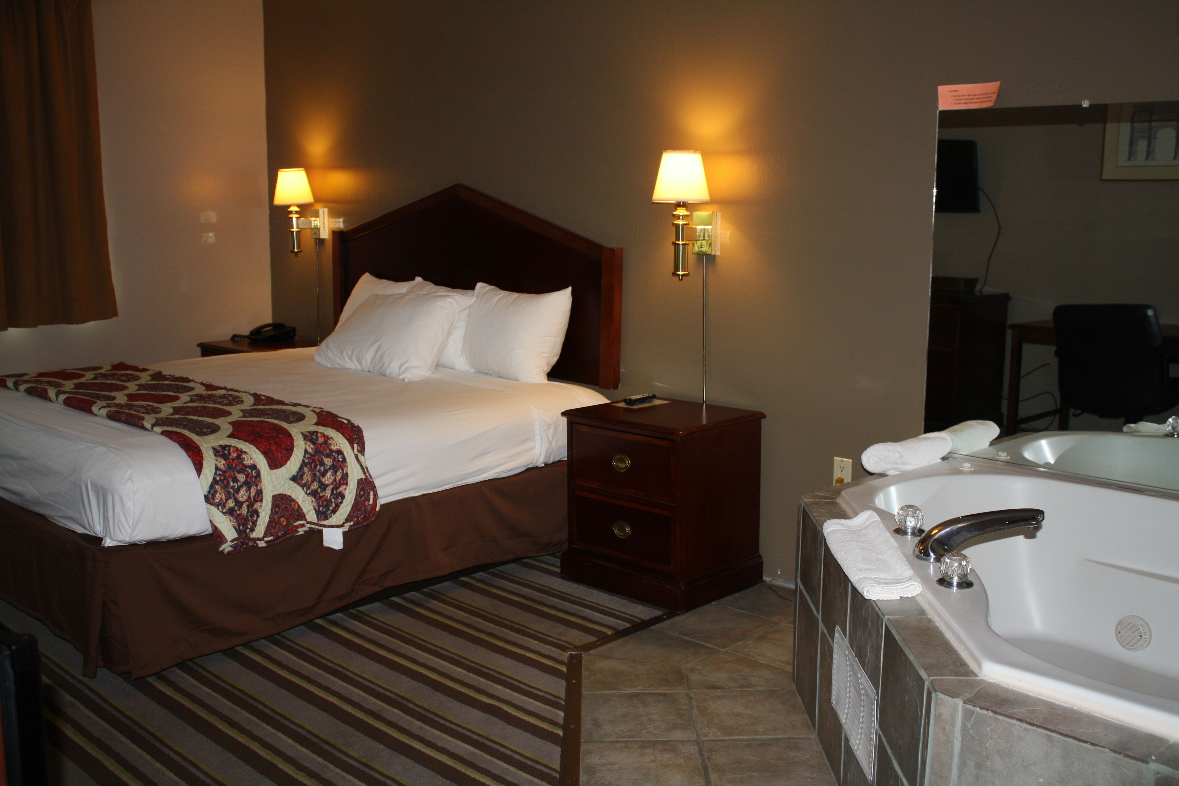 Quarto de hotel com uma cama grande, mesa de cabeceira, escrivaninha, luminária de mesa e uma banheira espaçosa, além de um espelho.