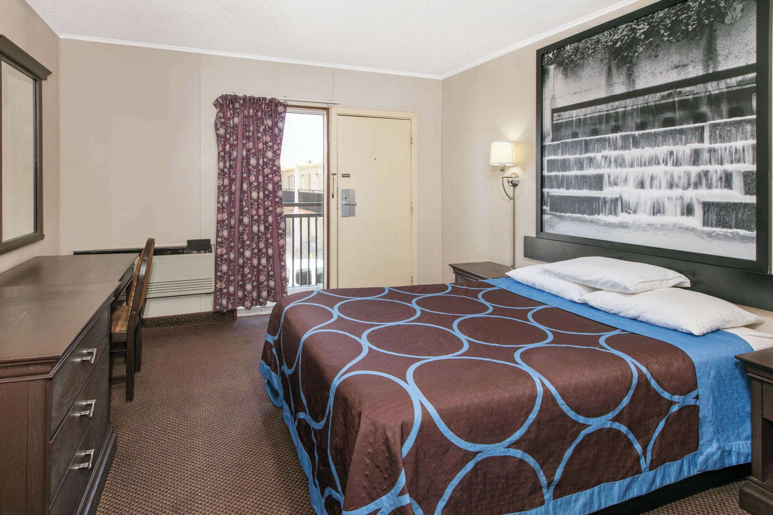 Quarto de hotel com cama, escrivaninha, cadeira, porta com vista e obra de arte na parede.