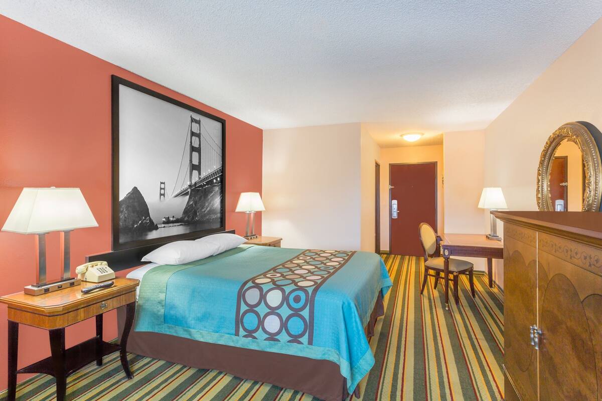 Quarto de hotel com uma cama, duas mesas de cabeceira com luminárias, uma escrivaninha com cadeira, um espelho grande e um quadro na parede.
